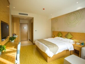 Quarto de hotel com cama de madeira, mesa de cabeceira com abajur, televisão presa na parede e planta no chão.