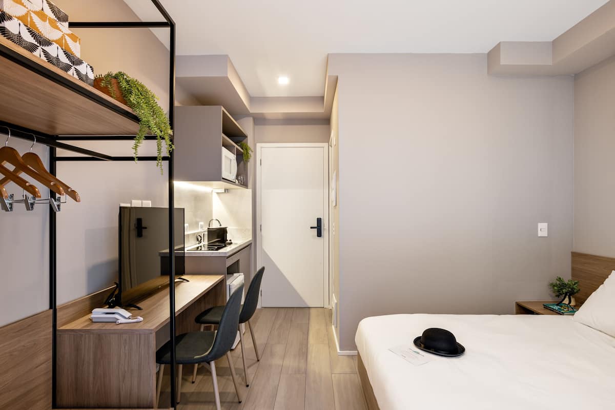 Quarto de hotel compacto com escrivaninha de madeira, cama, mesinha de cabeceira e guarda-roupa.