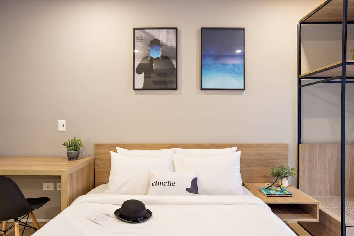 Quarto de hotel moderno com cabeceira de madeira, cama com roupa de cama branca, um chapéu preto, uma pequena mesa de trabalho com uma planta e duas obras de arte emolduradas na parede.
