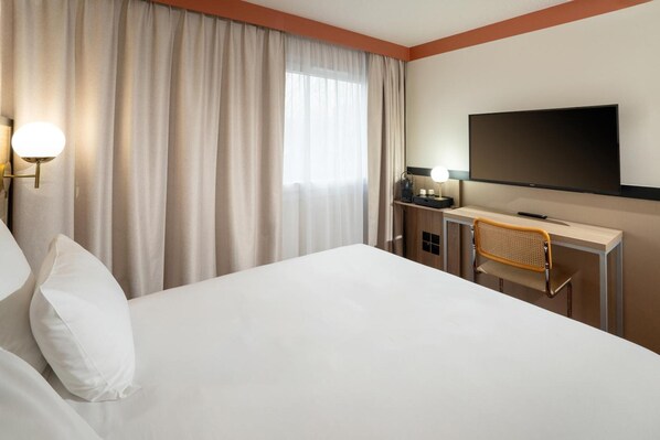 Quarto de hotel com uma cama grande, uma mesa com televisão de tela plana, uma cadeira e uma janela com cortinas.