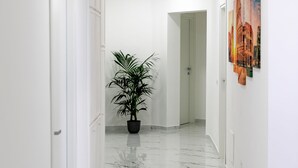 Corredor com paredes brancas, piso de mármore, uma planta em vaso e uma porta no final.