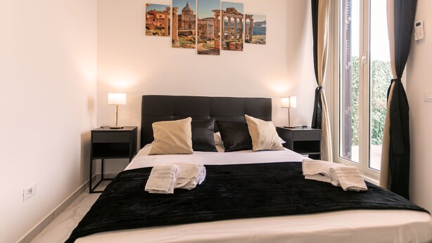 Cama bem arrumada com colcha preta e lençóis brancos, duas mesas de cabeceira com abajures e uma parede com uma série de quadros retratando arquitetura histórica.