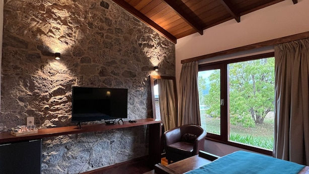 Quarto com parede de pedra, televisão, cama, cadeira e janela com cortinas.