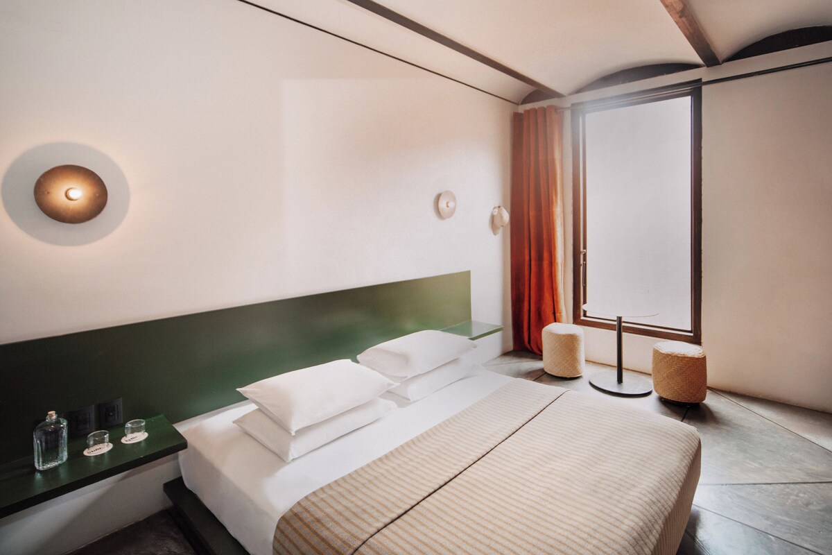 Quarto de hotel com cama, mesas de cabeceira, janela com cortinas e luminária fixa na parede.