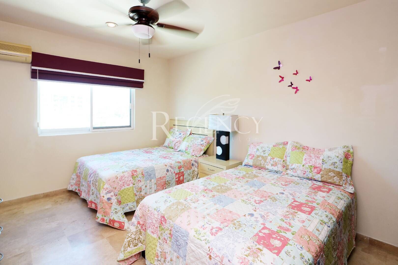 Quarto com duas camas, um ventilador de teto, janela com persianas e uma decoração de borboletas na parede.