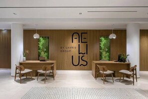 Uma área de recepção moderna com mesas e cadeiras de madeira, uma parede verde com folhagem e o logotipo de 'AE LAVRIS GROUP'.