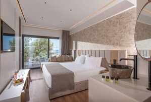 Quarto de hotel moderno com uma cama grande, banheiro com cuba de pedra, área de refeições com mesa e cadeiras, e vista para o exterior.
