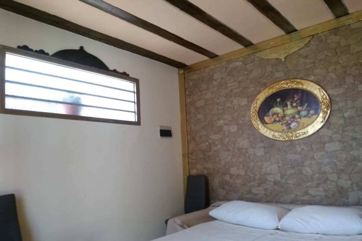 Quarto com parede de pedra, cama com roupa de cama branca, janela com persianas e um quadro na parede.