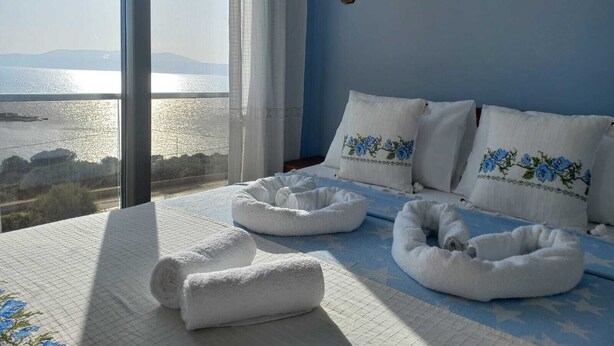 Quarto com uma grande janela com vista para um corpo de água, uma cama com roupa de cama branca e almofadas florais, e uma pequena prateleira de madeira com um objeto decorativo.
