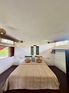 Quarto simples e limpo com uma cama, duas almofadas, janela com cortinas e luminária suspensa.