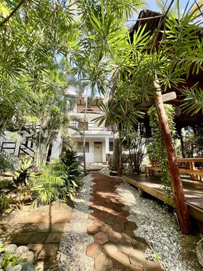 Uma trilha leva a um edifício rodeado por vegetação exuberante e uma construção com telhado de palha.