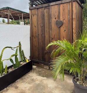 Um portão de madeira com um recorte em formato de coração, piso de concreto e vasos com plantas, incluindo uma palmeira.