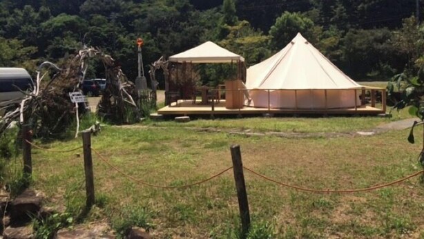 Alojamento tipo tenda com telhado plano, rodeado por uma área gramada e uma colina arborizada.