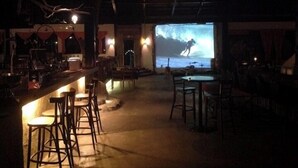 Um bar interior com uma grande tela exibindo um vídeo de surf, banquetas e mesas.