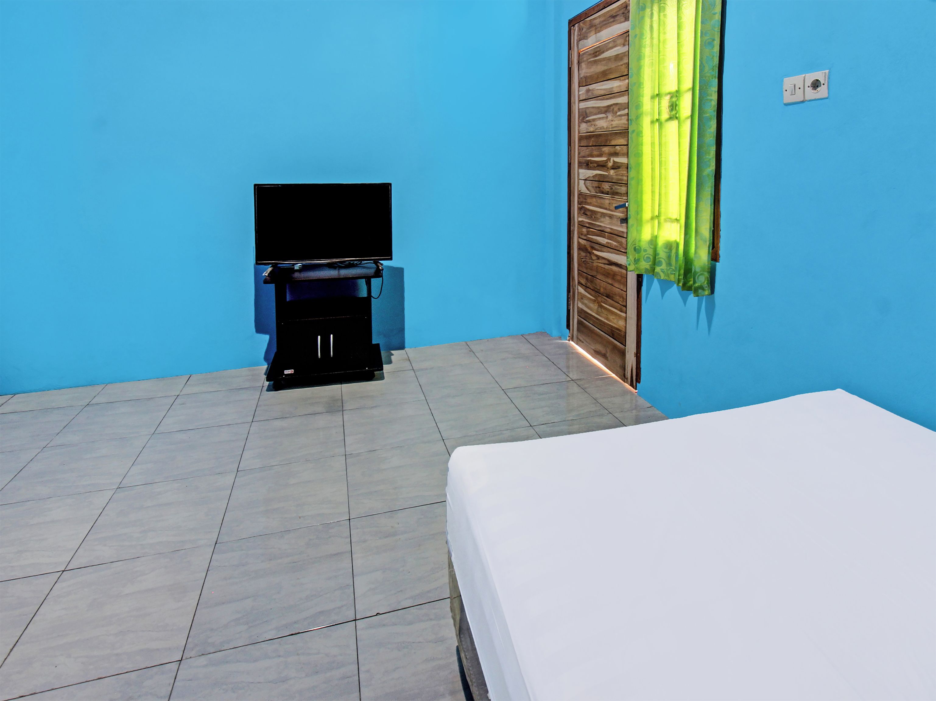 Um quarto com uma cama, uma televisão sobre um suporte e uma porta de madeira com cortina verde.