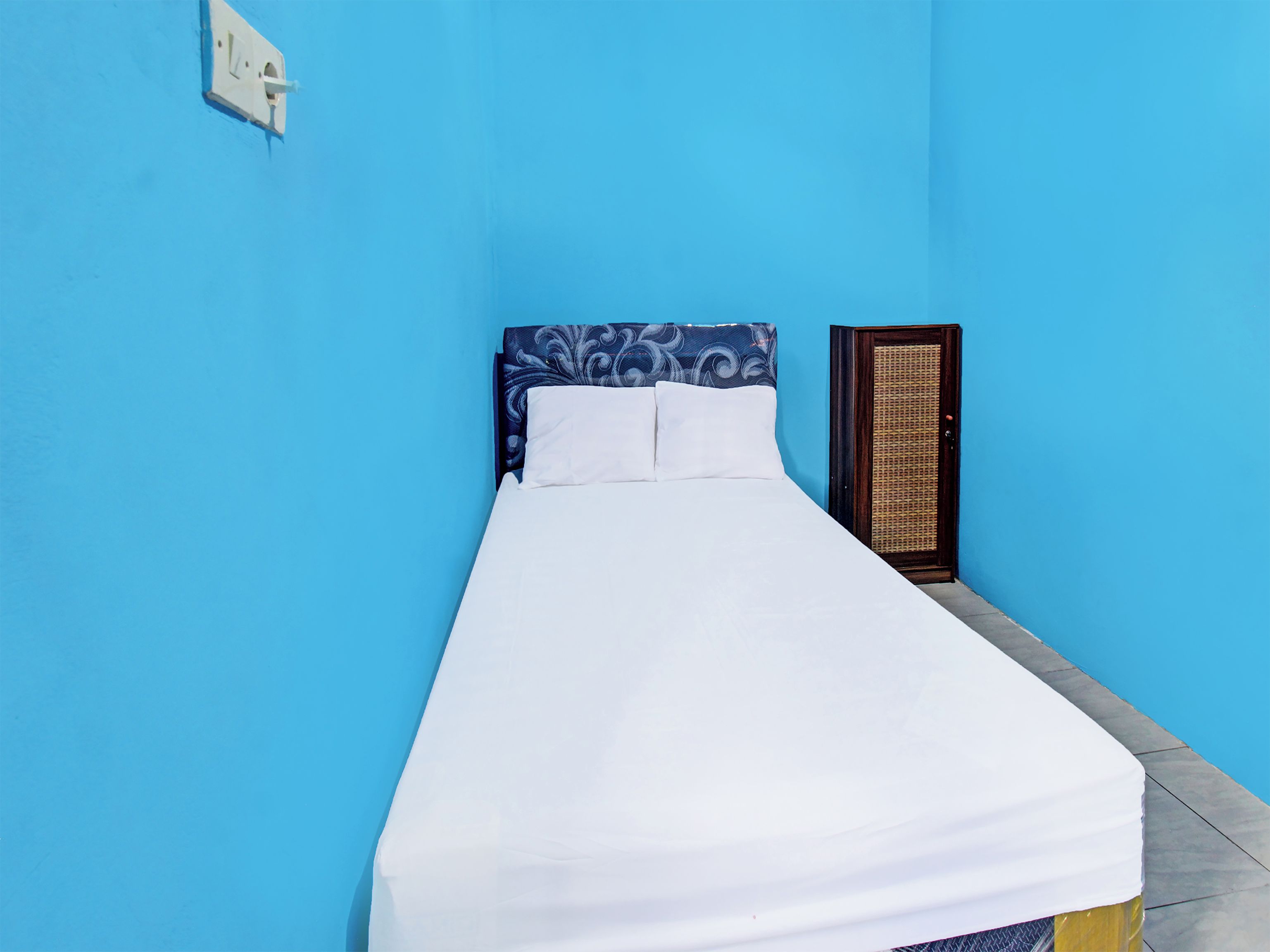 Uma cama de solteiro com colchão branco e cabeceira azul estampada, uma porta de vime e uma parede azul.