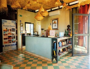 Uma área de recepção com piso xadrez, balcão de atendimento, luminárias de vime suspensas, quadros emoldurados e uma vitrine de vidro.