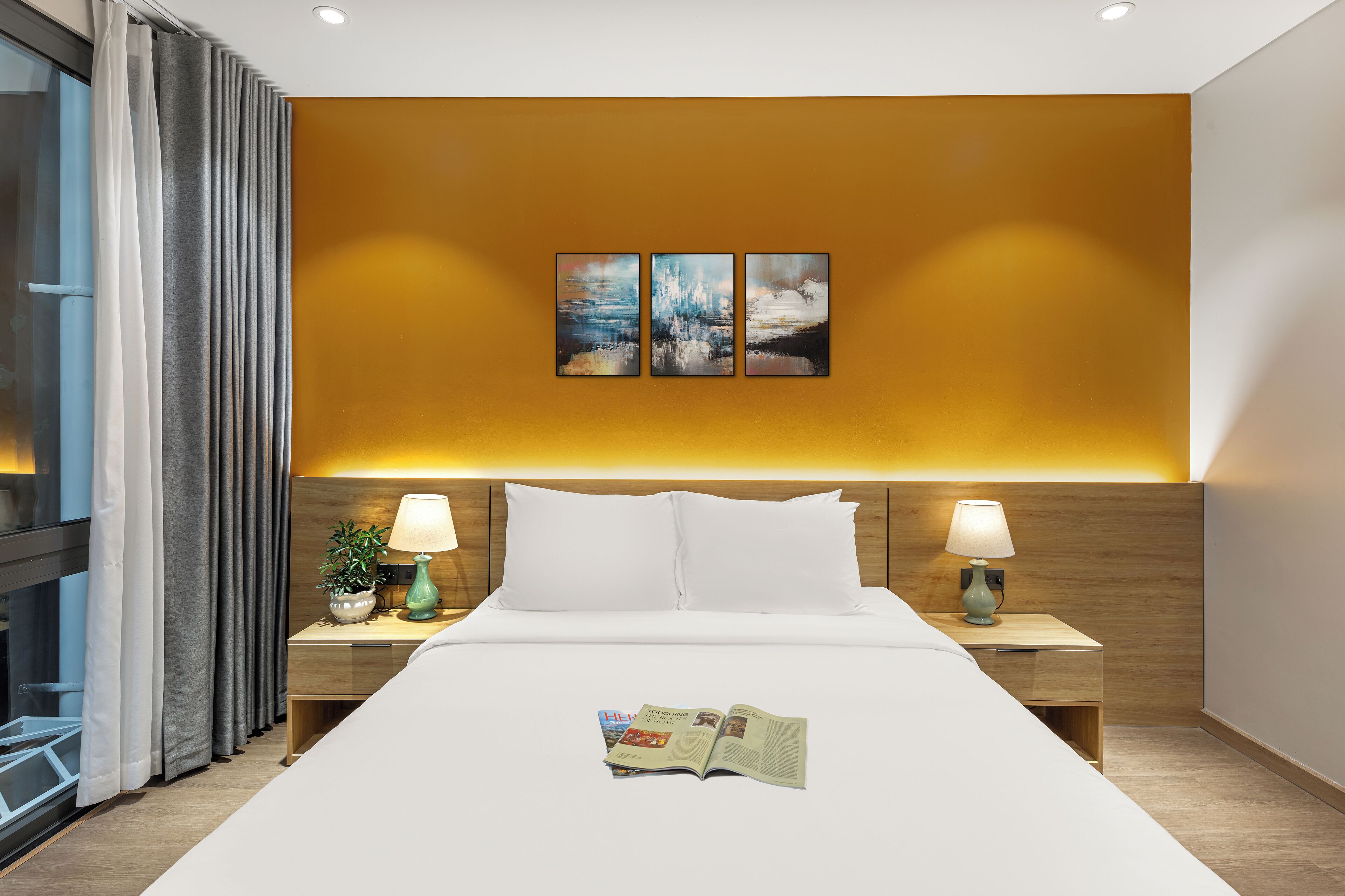 Quarto de hotel com uma cama grande, duas mesas de cabeceira com luminárias, um criado-mudo com uma planta e uma obra de arte na parede acima da cama.