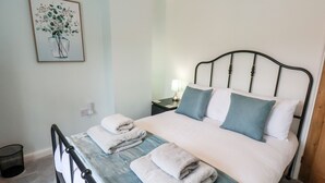 Uma cama bem arrumada com duas almofadas, uma mesinha de cabeceira com abajur e um quadro na parede.