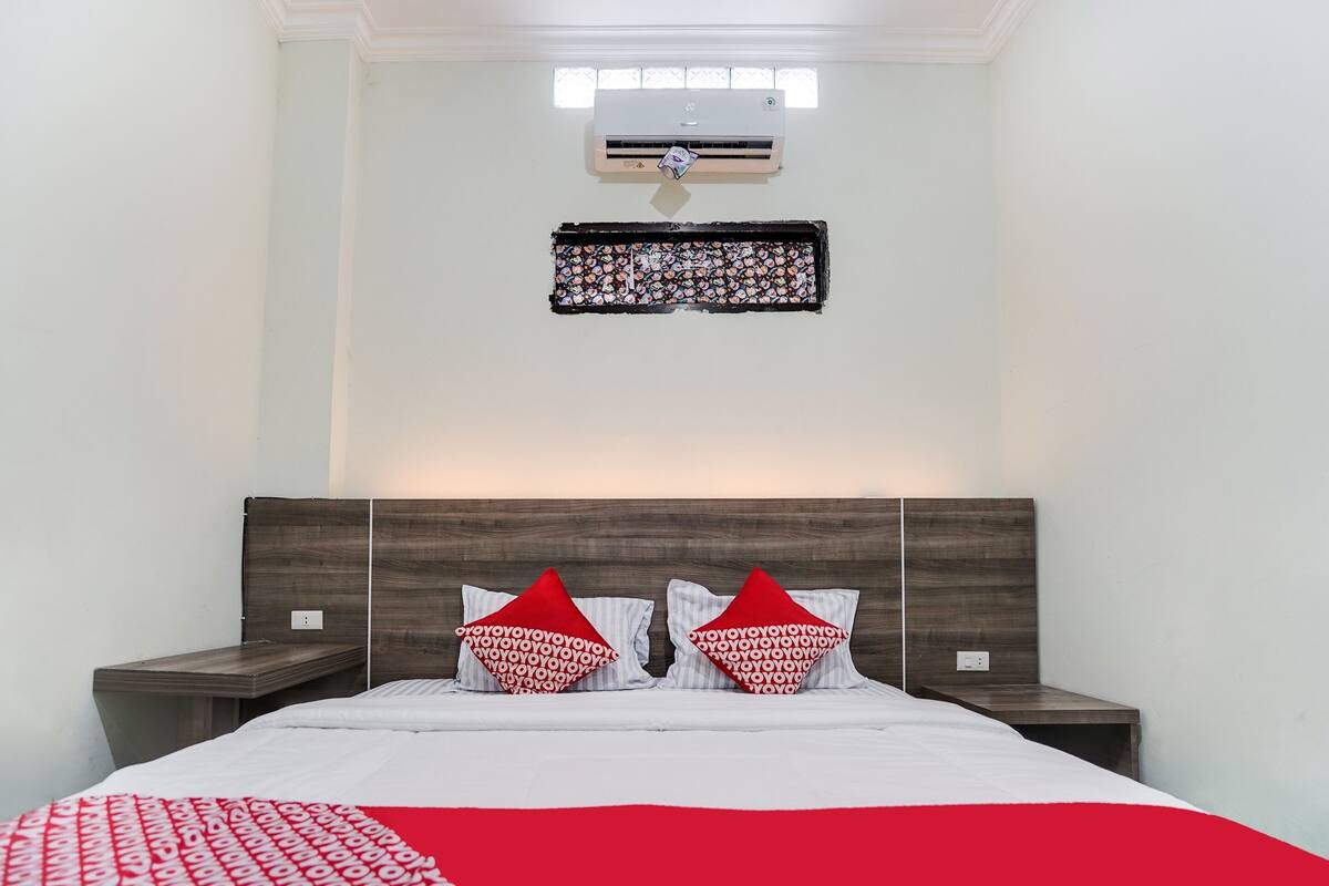 Quarto de hotel bem organizado, com uma cama, duas mesas de cabeceira, ar condicionado fixado na parede e um pequeno objeto decorativo sobre a cama.