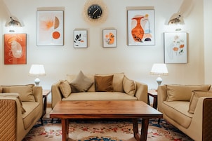 Sala de estar com sofá bege, duas poltronas, mesa de centro de madeira e tapete estampado. Quadros emoldurados na parede e duas luminárias de parede.
