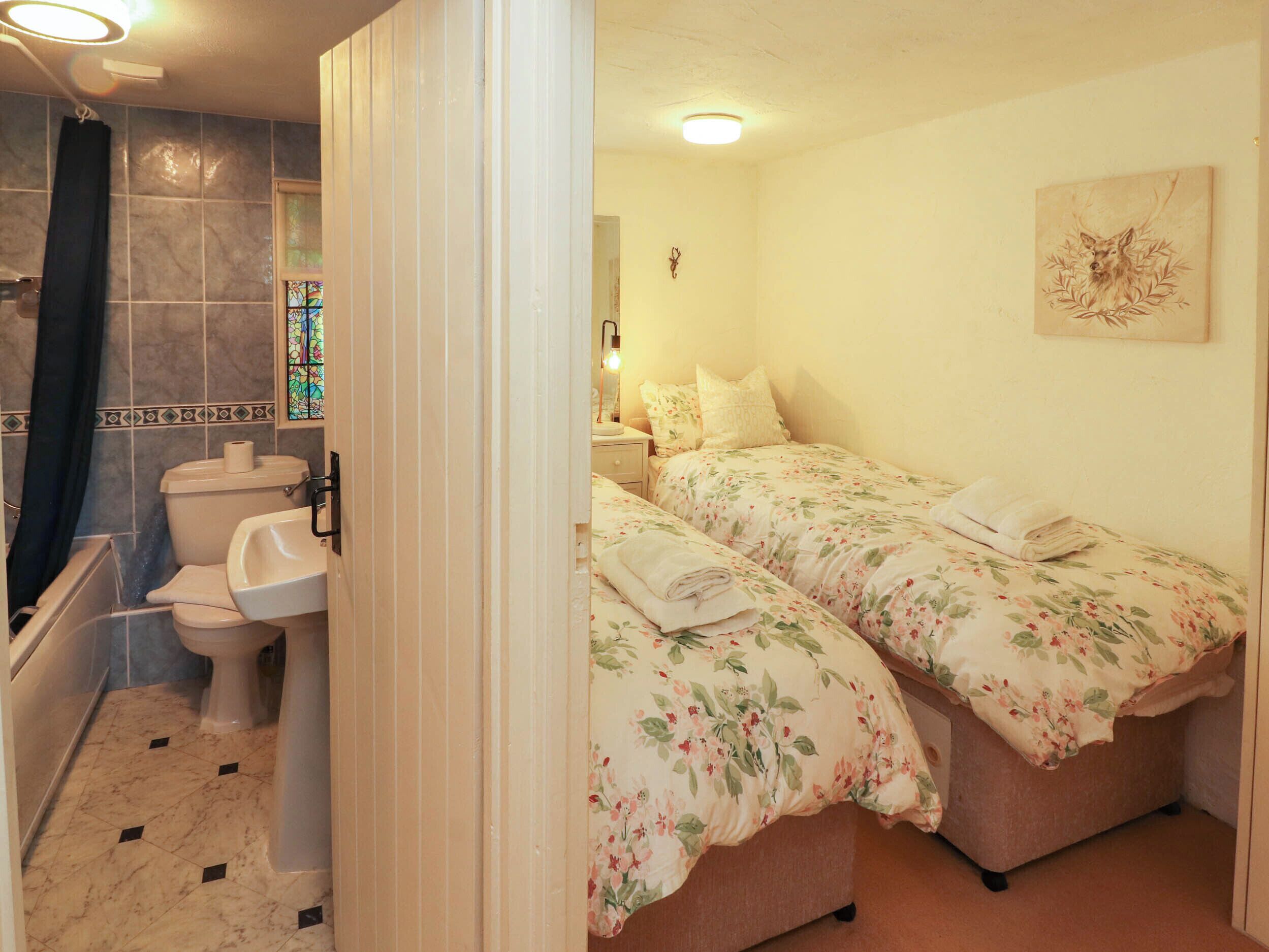 Casa de banho com chuveiro, sanita e lavatório. Existem duas camas com roupa de cama floral e um quadro na parede.