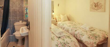 Casa de banho com chuveiro, sanita e lavatório. Existem duas camas com roupa de cama floral e um quadro na parede.