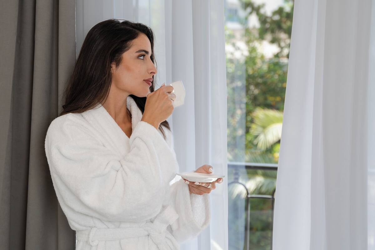 Uma pessoa em um roupão branco segurando uma xícara branca perto de uma janela com cortinas leves.