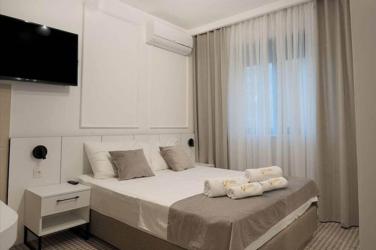 Quarto de hotel com uma cama, televisão fixada na parede, mesa de cabeceira e janela com cortinas.