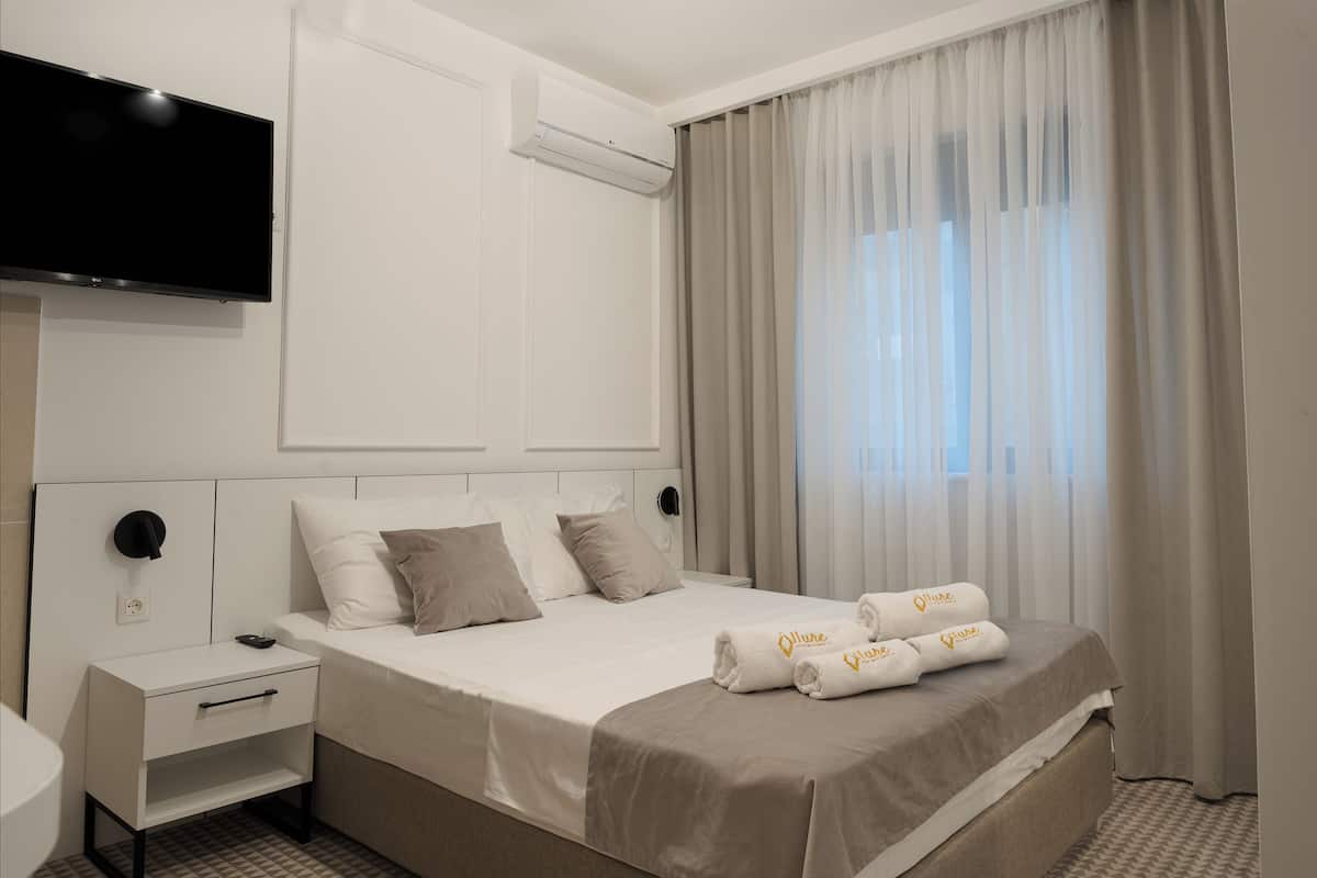 Cama bem arrumada com colcha branca e bege, dois travesseiros e duas toalhas enroladas com um logotipo. Há uma TV de tela plana na parede acima da cama, uma mesinha de cabeceira com gaveta e uma janela com cortinas leves.