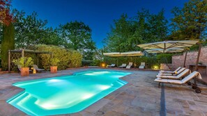 Uma piscina com espreguiçadeiras e guarda-sóis, cercada por árvores e uma pérgola.