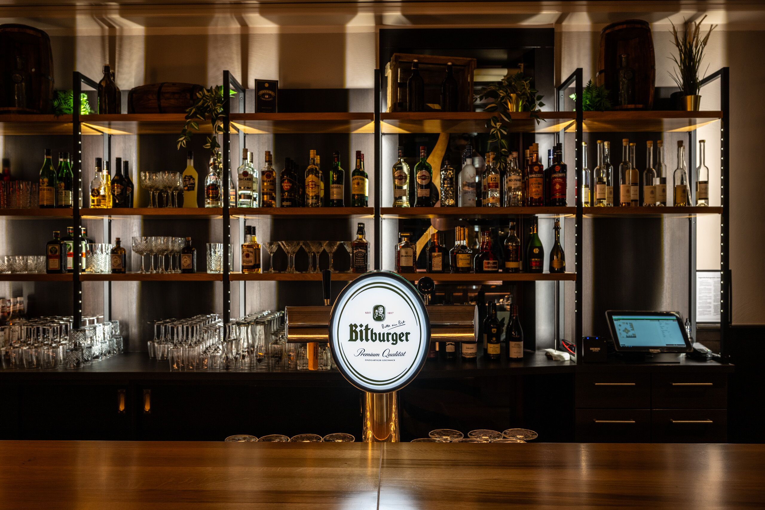 Uma área de bar com uma torneira de cerveja Bitburger, prateleiras com diversas garrafas de bebidas alcoólicas e um monitor de computador em um balcão.
