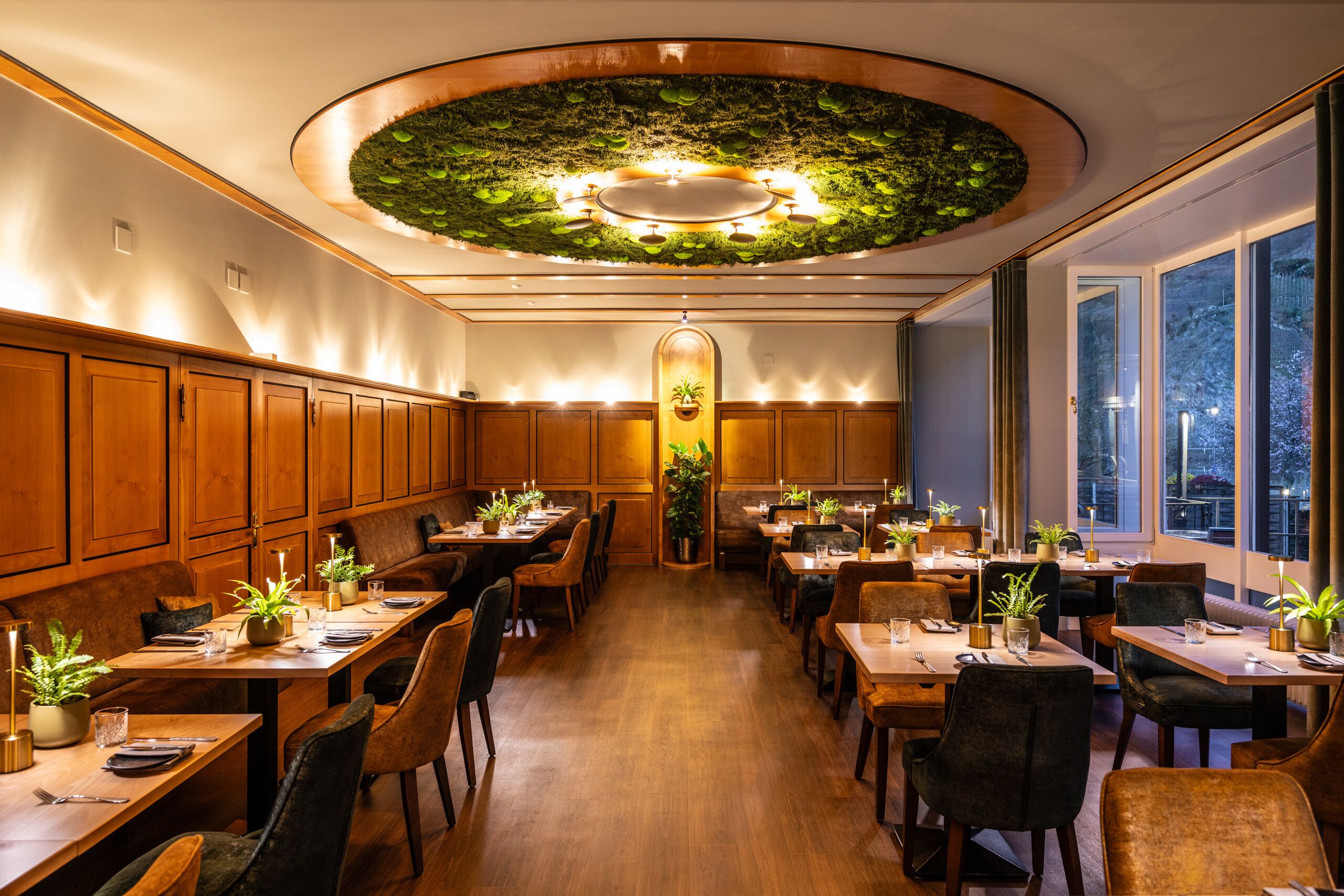 Restaurante com painéis de madeira, mesas com plantas em vasos e teto com design de folhagem verde.