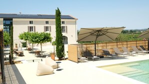Uma área de piscina no terraço, com espreguiçadeiras, um grande guarda-sol e um edifício com telhado de azulejos.