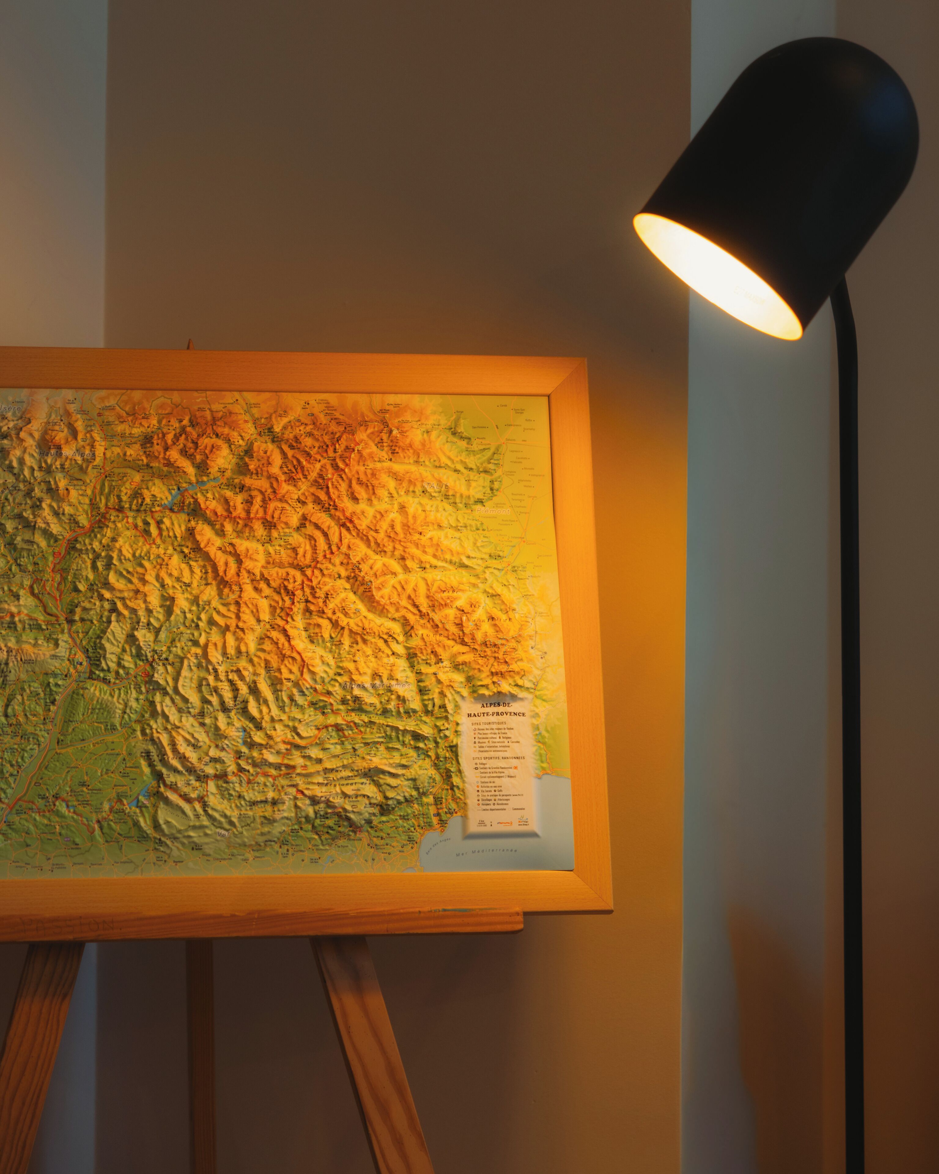Um mapa topográfico emoldurado, um abajur de mesa preto e um cavalete de madeira.