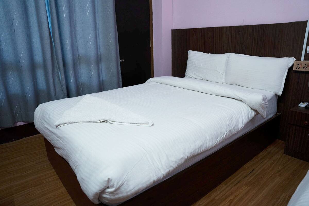 Cama bem arrumada com roupa de cama branca em quarto com piso de madeira e cabeceira escura.