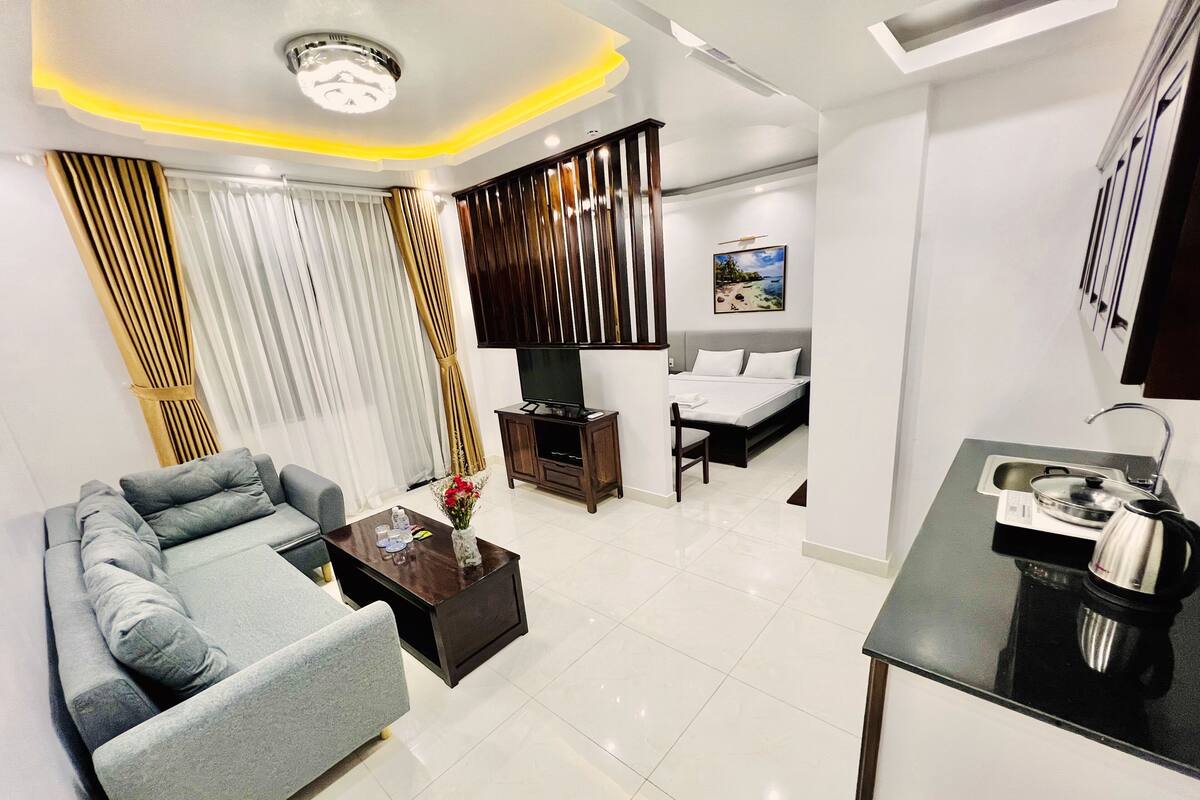 Uma sala de estar moderna com sofá, mesa de centro com um vaso de flores, televisão, cama e área de cozinha com chaleira e pia.