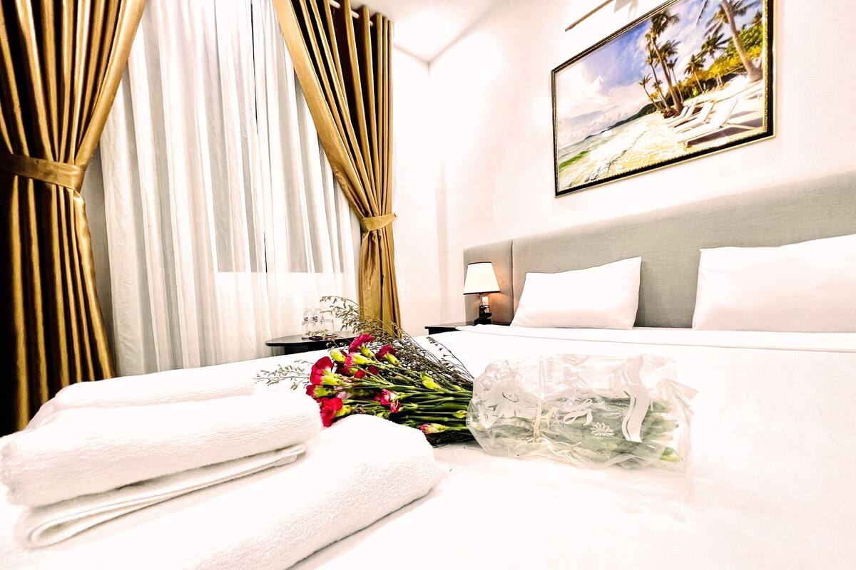 Quarto de hotel com cama, cortinas, mesa de cabeceira, abajur, um arranjo de flores e um quadro na parede.
