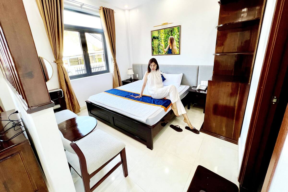 Quarto de hotel com cama, escrivaninha, cadeira, mesa de cabeceira, abajur e um quadro na parede.
