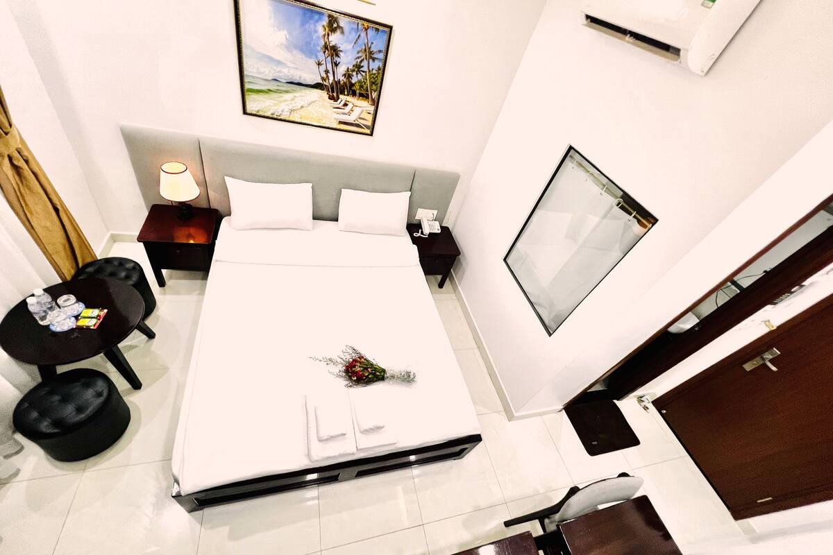 Quarto de hotel com cama, escrivaninha com cadeira, mesa de cabeceira, luminária, quadro na parede e janela.