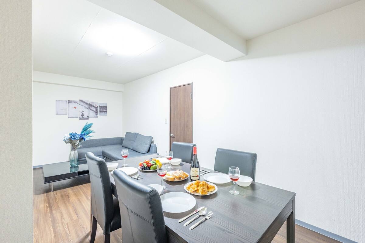 Uma sala de jantar com uma mesa de madeira escura, cadeiras estofadas cinzentas e um sofá cinza. Há pratos de comida, uma garrafa de vinho e um vaso com flores sobre a mesa.