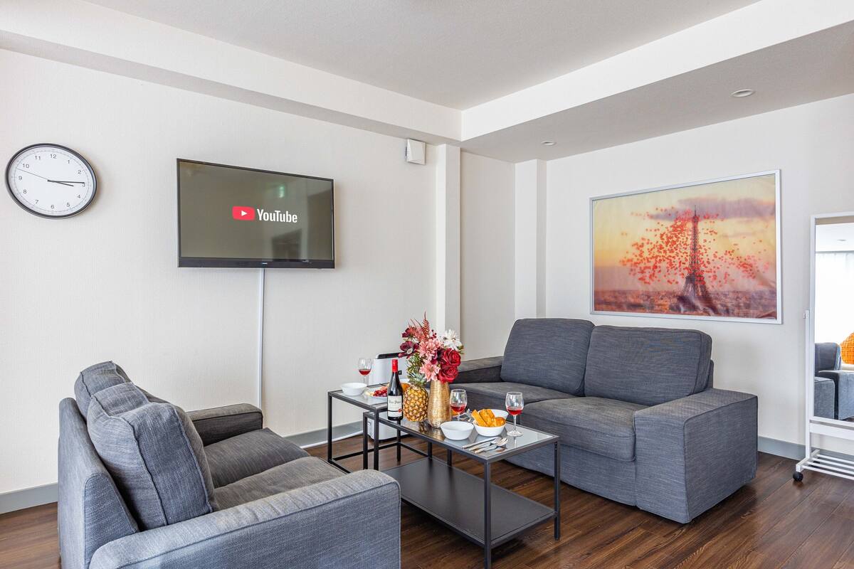 Uma sala de estar moderna com televisão de tela plana, um relógio, um sofá, uma mesa de centro com um vaso de flores e um quadro da Torre Eiffel.