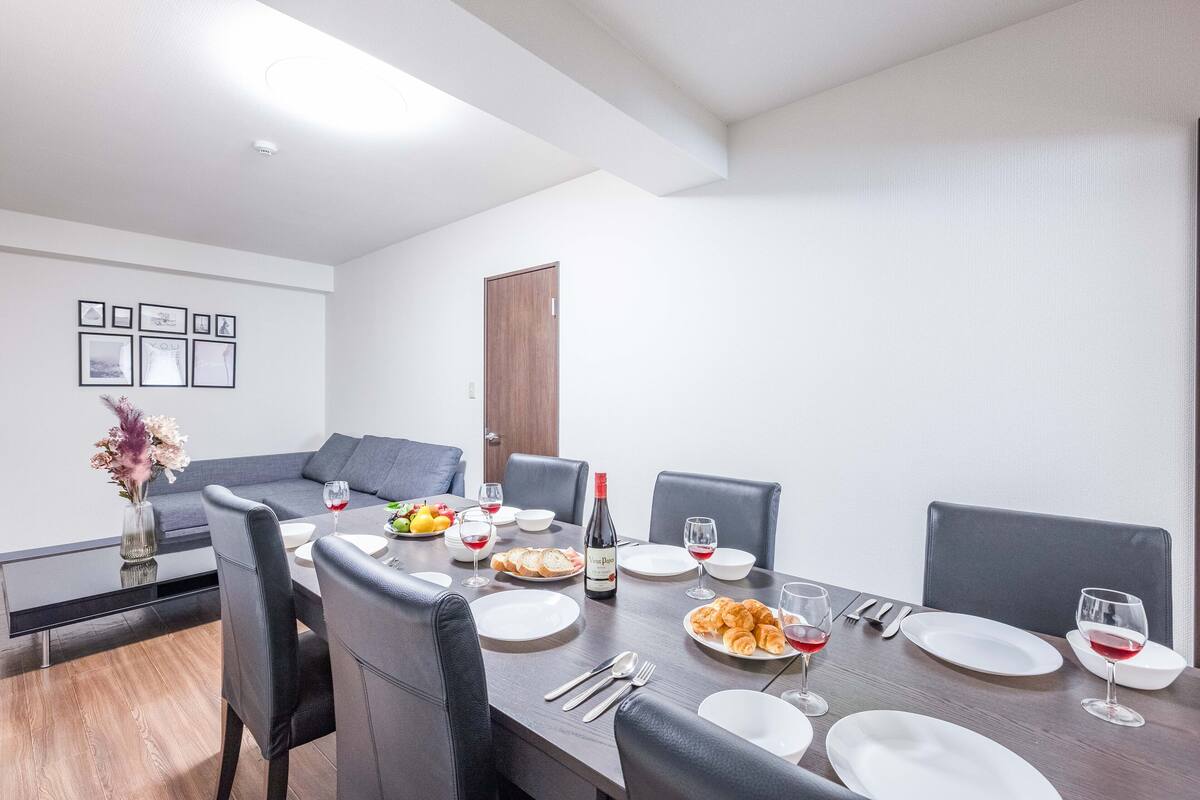 Uma sala de jantar moderna com uma mesa comprida posta para quatro pessoas, rodeada por cadeiras de couro escuro. Há pratos, copos e uma garrafa de vinho sobre a mesa.