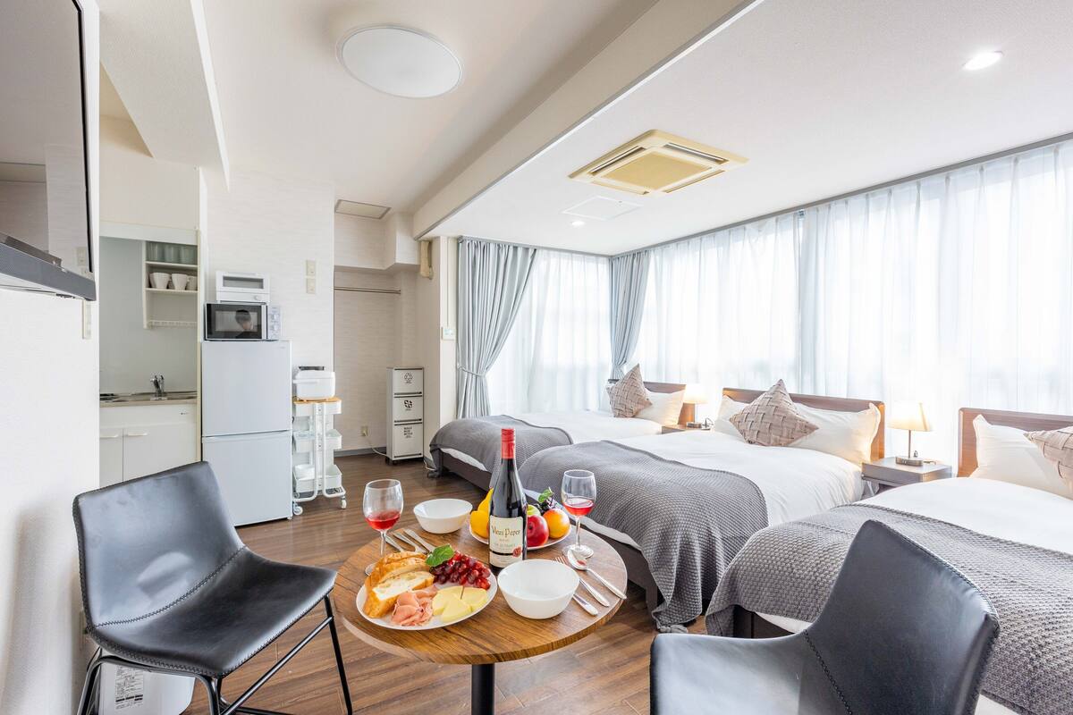 Quarto de hotel com mesa de jantar posta com comida e bebidas, duas camas, cozinha compacta e uma janela grande com cortinas.