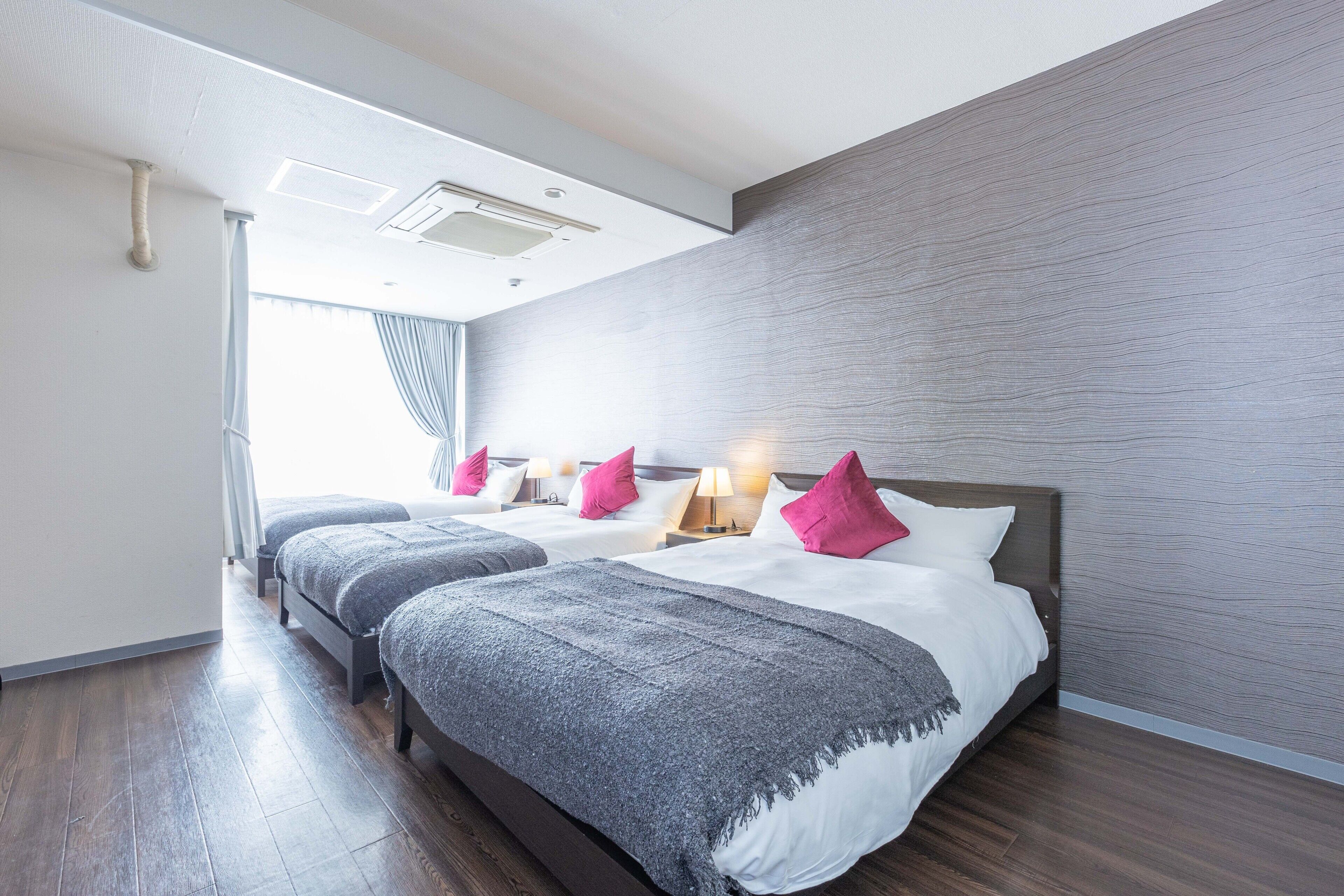 Quarto de hotel moderno com duas camas, uma janela grande com cortinas e piso de madeira.