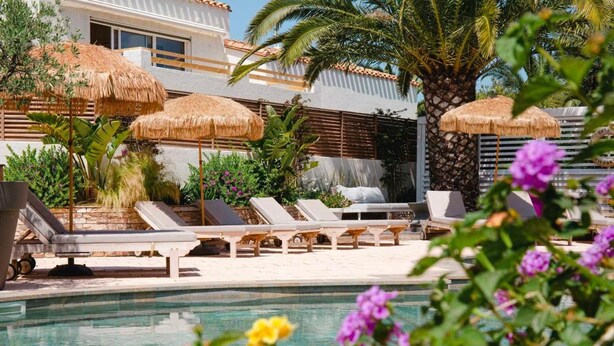 Uma área de piscina com espreguiçadeiras, guarda-sóis de palha e plantas tropicais.