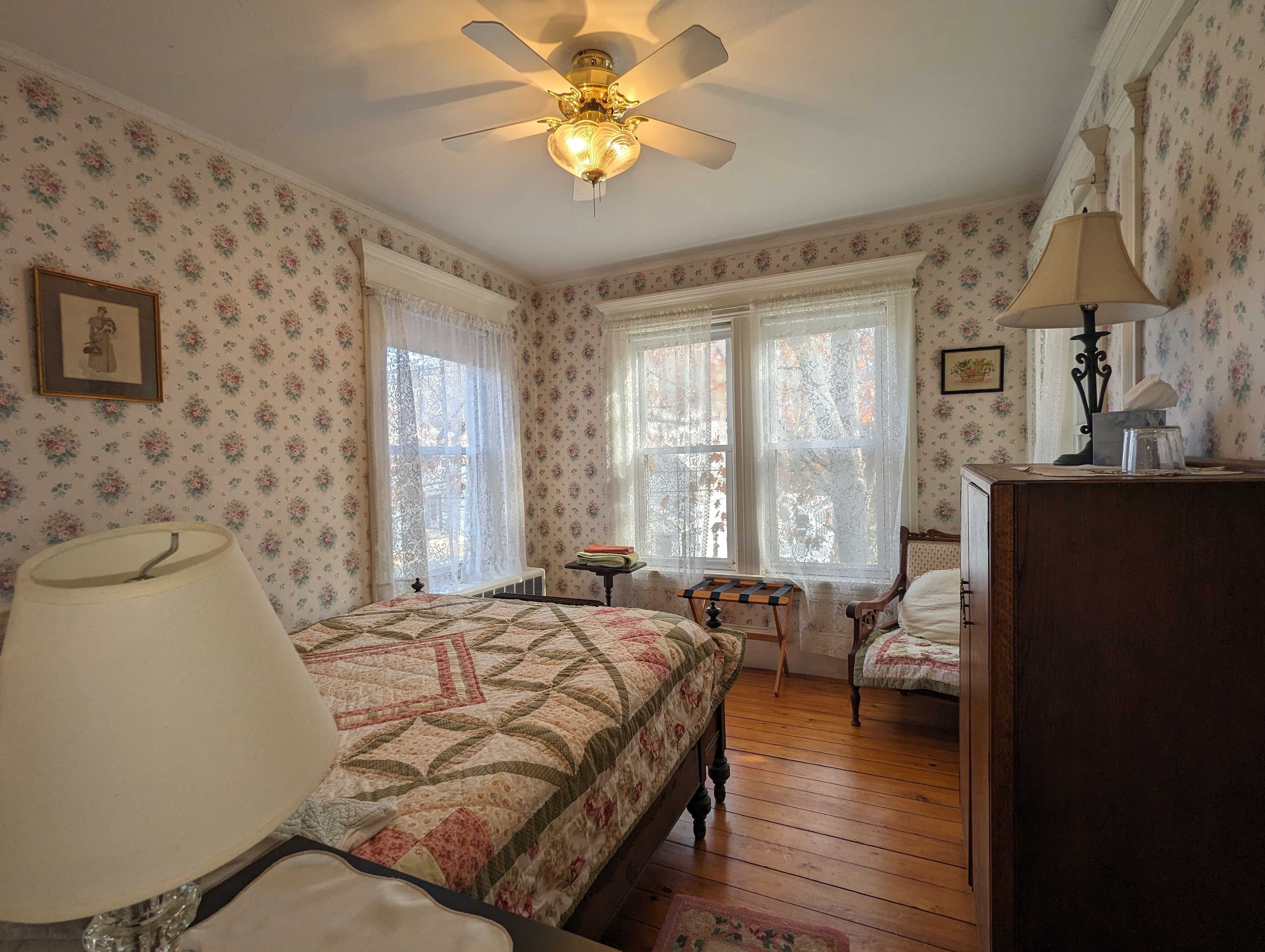 Quarto com papel de parede floral, cama com colcha, escrivaninha com abajur e ventoinha no teto.