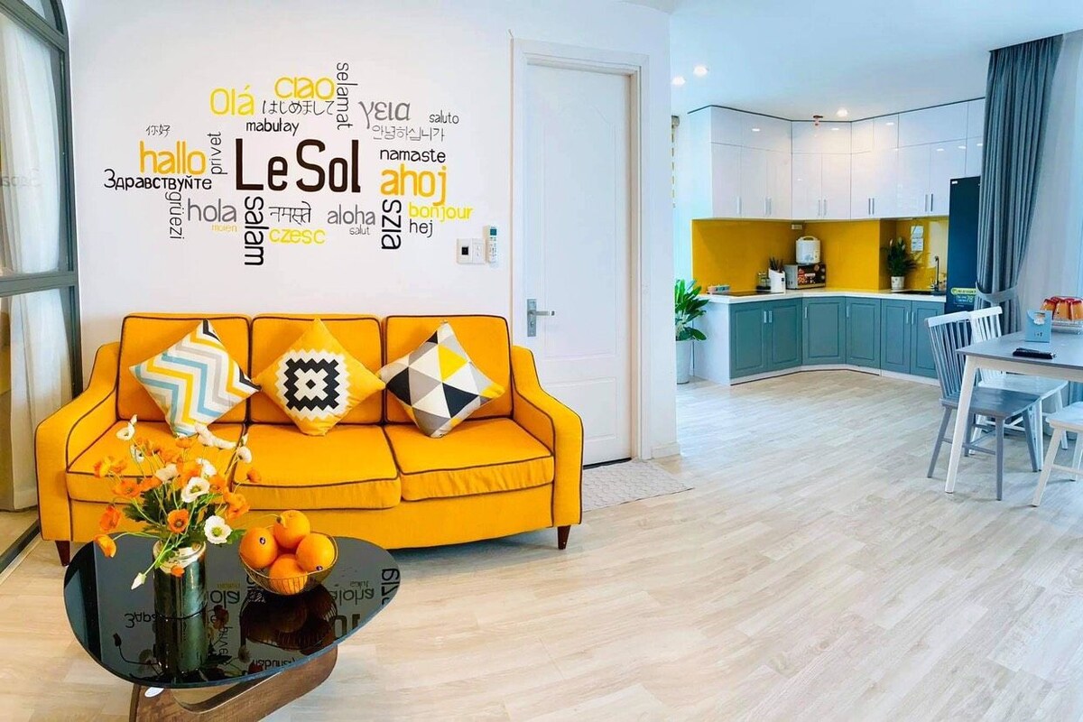 Uma sala de estar moderna com um sofá amarelo, uma mesa de centro de vidro com flores e uma obra de arte com palavras na parede.