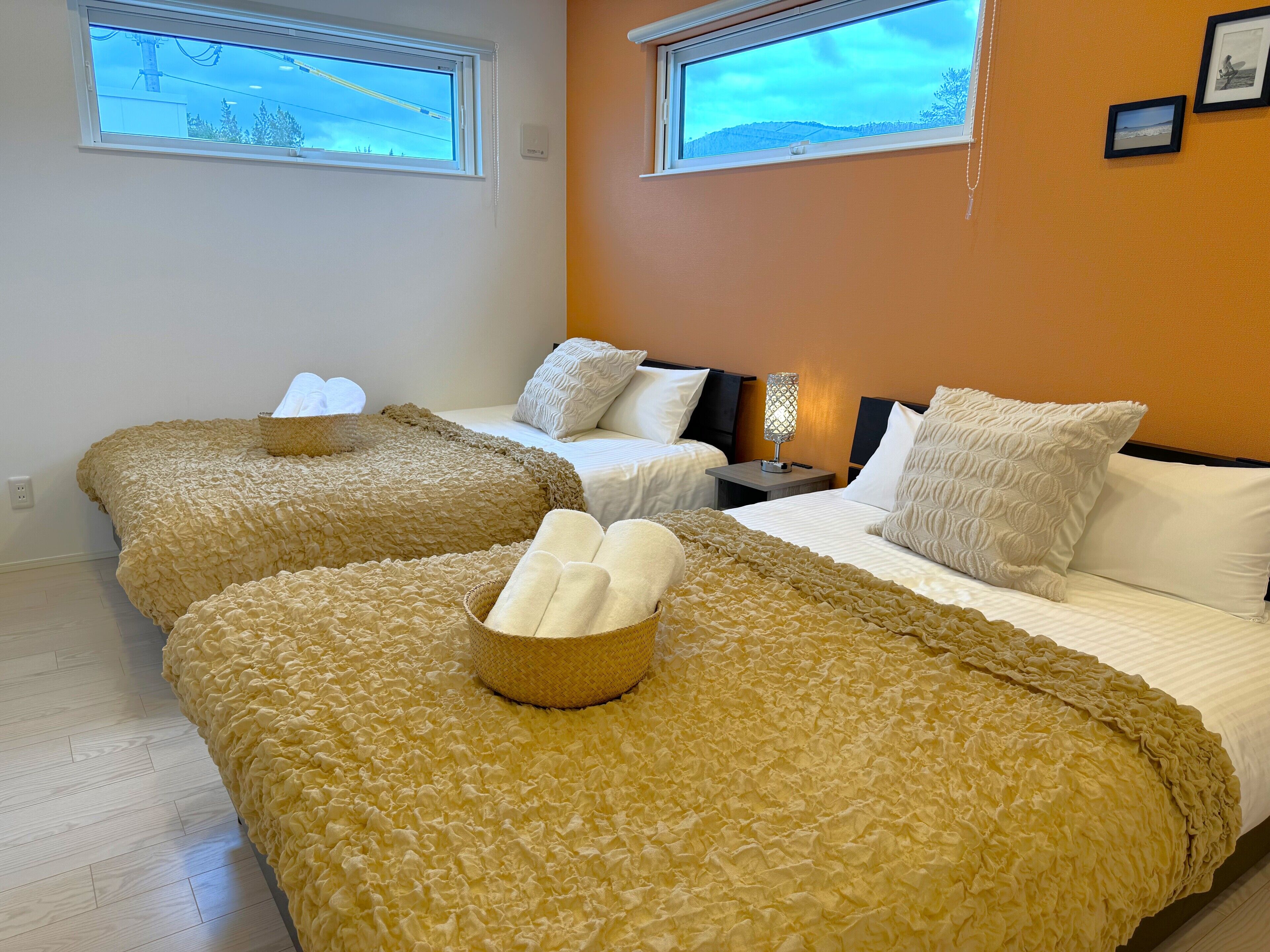 Quarto de hotel com duas camas, cada uma com um cobertor bege texturizado e um travesseiro branco. Há uma mesinha de cabeceira com uma luminária entre as camas.