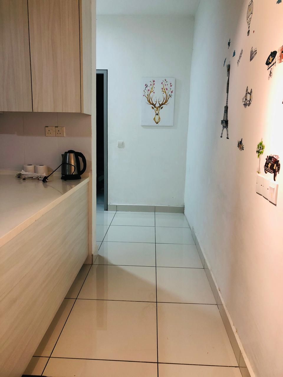 Uma cozinha moderna com armários de madeira, uma chaleira e uma cafeteira na bancada. Um corredor com piso ladrilhado leva a um quarto com uma porta.